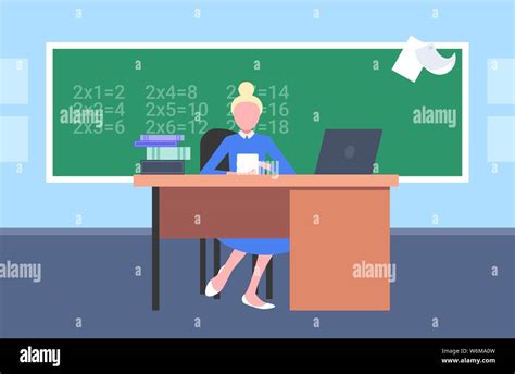 Female Teacher Sitting At Desk In Front Of Chalkboard Using Laptop Math
