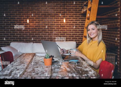 Attractive Young Blonde Woman Sitting At Table In Her Modern Loft