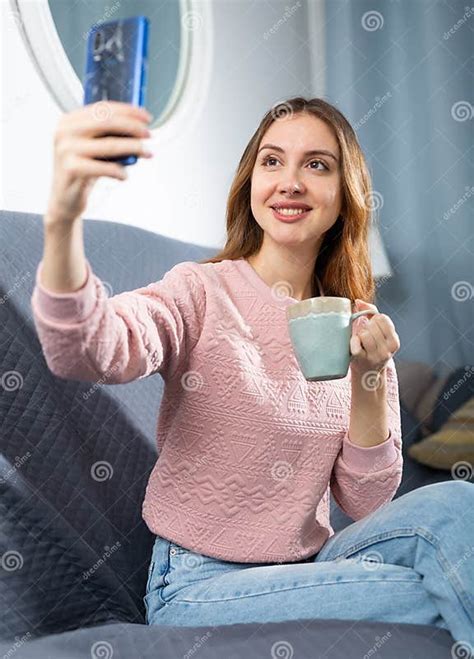 Young Woman Taking Selfie While Sitting On Sofa With Mug Of Tea Stock