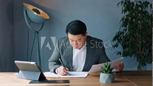 Thoughtful сhinese executive meticulously examining documents in office environment. Attentive young businessman filling out paperwork and completing form or application with focused attention.