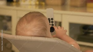 Baby boy sits in rocking chair and holds remote control from TV digital media player. Child watches cartoons on TV. High quality 4k footage
