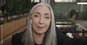 Close up of elegant middle aged businesswoman in earpods having videocall on smartphone. Camera view of beautiful old woman communicating, using video app of her phone while sitting in cafe.