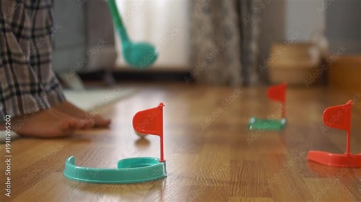A focused shot on the mini golf toy set on the floor where a child’s hands and feet appear as they align the ball and take a swing with a plastic club.