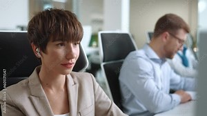 Pretty Caucasian woman manager with suit using earpods in office. Young busy female sitting in front of computer while listening music. In blurred background men working on PC. Technology concept.