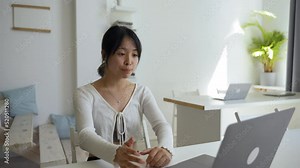 Cheerful Asian woman waves hello on video call with friend via modern laptop. Young employee takes break at work for online communication closeup