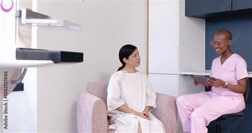 Woman clinician in scrubs consulting tablet after mammogram while patient in gown listening