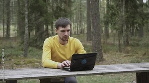 Young brunet in yellow longsleeve looking at laptop screen in forest. Guy is surprised, greatly rejoices at victory in game, gambling sports bet. Happy man waving arms, banging fist, clapping hands.