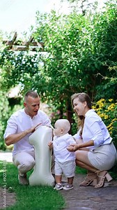 Cute little blond baby boy stands near his parents. Smiling dad shows number one balloon to his kid. First child's birthday. Vertical video.