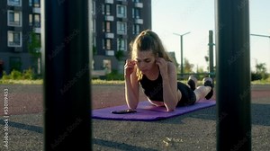 pretty slender young woman puts headphones in her ears after or before exercising while lying on gymnastic mat early in the morning outdoors on sports field in yard of her residential complex.