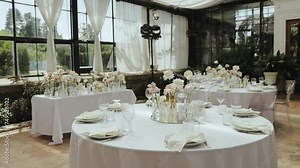 Pastel tender bouquets of roses in glass vase on festive rounded tables at the wedding. Large windows with transparent curtains on the background. Wine glasses and plates, wedding in orangery.