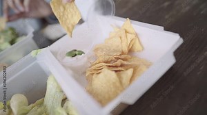 Close up female hand pick up tortilla chips and dip on sour cream sauce, appetizers menu, take away delivery food, delivery box container, snacking before having main course, Mexican food culture