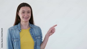 Smiling confident girl in braces on white background, points finger on space for text. Orthodontic clinic advertisement. Dental treatment by brackets for adult person. Happy woman with braces on teeth