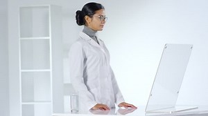 Medium shot of Asian female scientist in lab coat swiping on futuristic PC with transparent monitor while standing at hi-tech desk. Video suitable for adding AR graphics