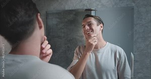 Young clean-shaven man carefully examines himself in bathroom mirror after getting ready morning. Millennial admires reflection in full-length mirror. Man pleased with way looks and smiling in mirror.