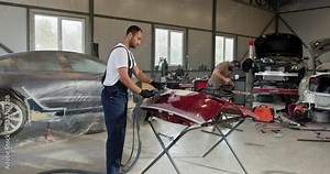 Professional auto mechanic worker polishing a red car bumper using a power grinder at an automobile repair and renew service station. Mechanic using grinding unit at automobile repair service station.