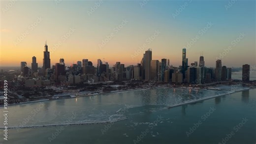 Drone view of downtown Chicago skyline at sunset during winter. Ice formations float on the surface of Lake Michigan as subzero temperatures create frozen patterns along the shoreline