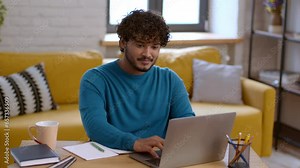 Positive indian guy working distantly at home office interior, typing and editing report on laptop, enjoying remote job