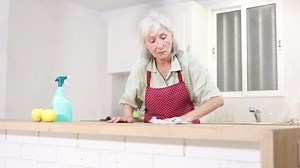 Senior woman in apron washing top of kitchen table during cleanup in apartment.