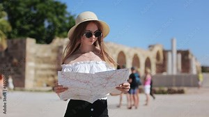 Young woman in straw hat and sunglasses holding paper map and checking direction while walking at resort city. Summertime, vacation and people concept.