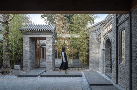A Woman Walking Through An Outdoor Courtyard With Stone Walls And Trees In The Back Ground