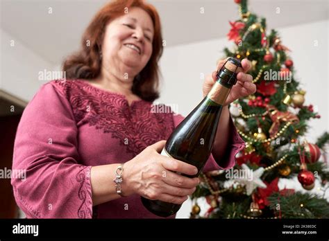 Latin Mature Woman Smiling While Opening A Bottle Of Champagne Or Cider With A Christmas Tree In