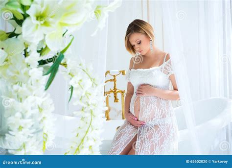 Pregnant Girl In A White Lace Dress Sitting In The Interior With Flowers And Hugs Belly Stock