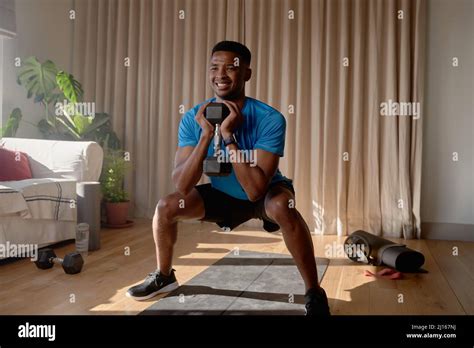 African American Male Squatting In Lounge Holding Dumbbell Weight Working Out At Home Stock
