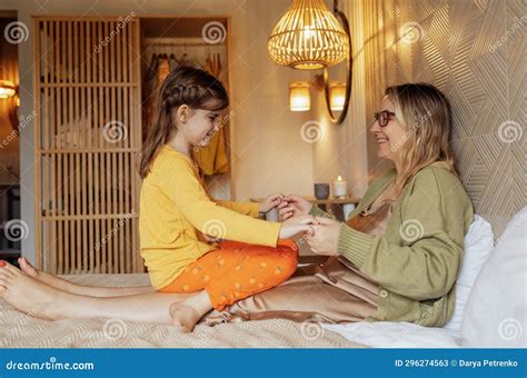 An Attractive Blonde Mom With Glasses And Her Cute Daughter Are Sitting On The Bed And Smiling
