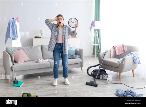 Shocked Young Lady With Clock Standing In Messy Room After Party Too Late To Clean Apartment