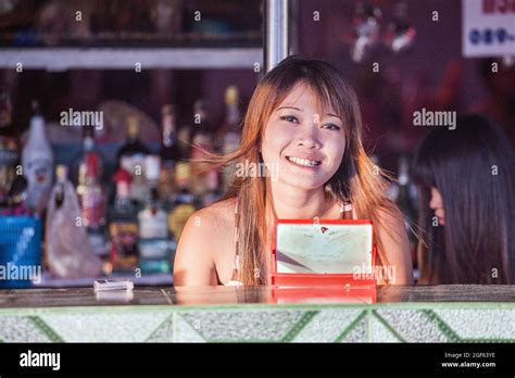 Pretty Thai Bar Girl Sits Smiling At Counter With Vanity Mirror In Front Of Her And Sun On Face