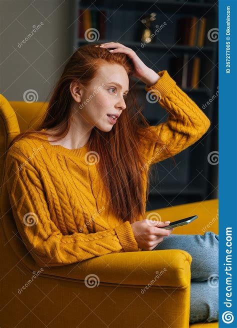 Vertical Portrait Of Attractive Redhead Woman Using Mobile Phone Sitting In Yellow Armchair