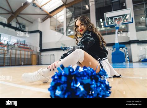 Brunette Curly Haired Cheerleader In Black And White Uniform And Jacket Sitting On Basketball