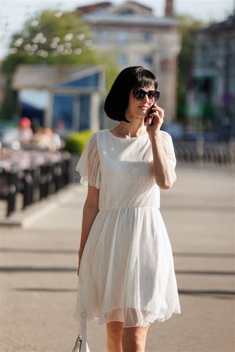 Romantic Young Brunette Woman With Short Hair Wears Mini White Dress And Sunglasses Talking On